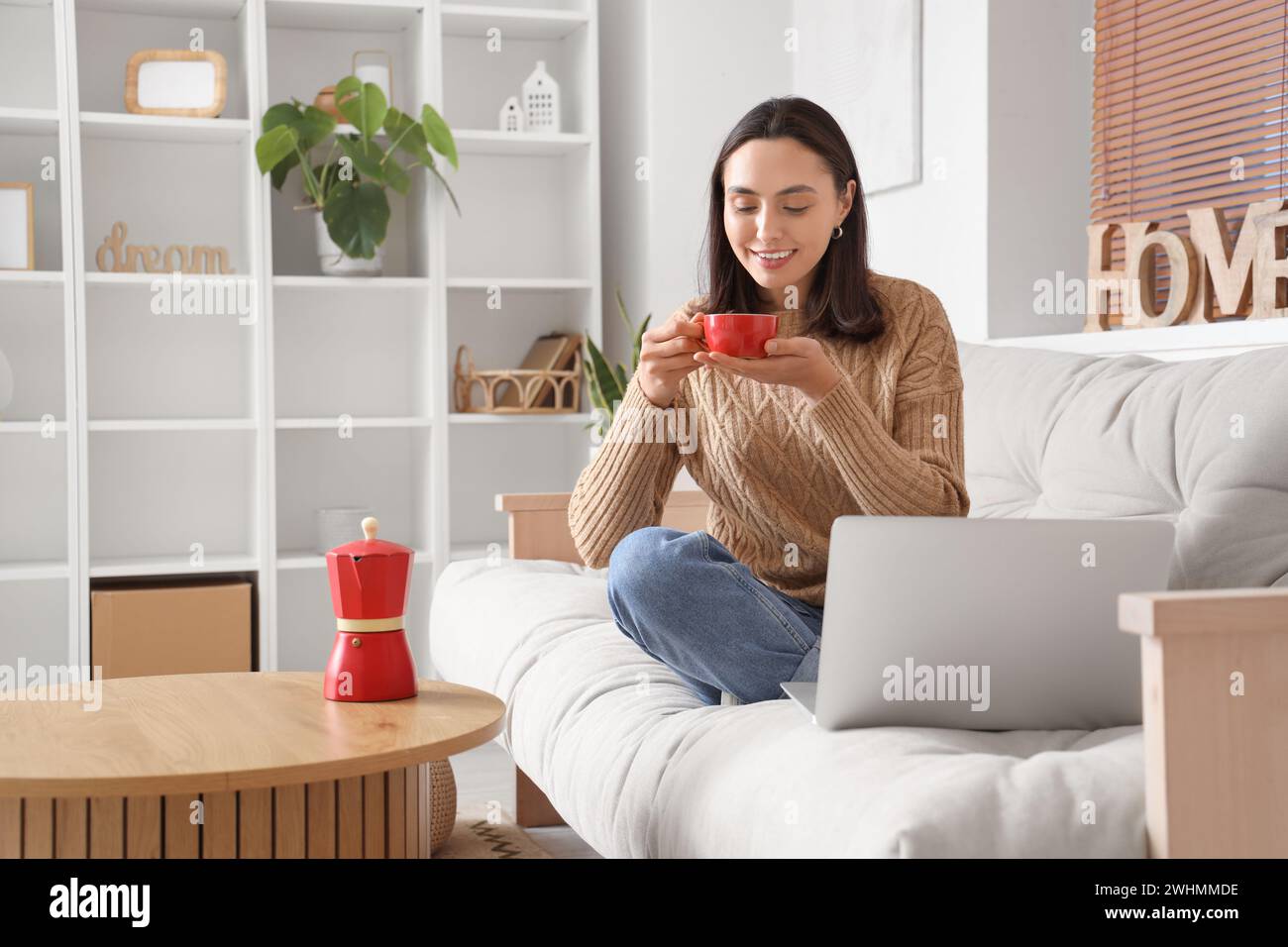 Pretty young woman enjoying coffee break at home with laptop on sofa ...