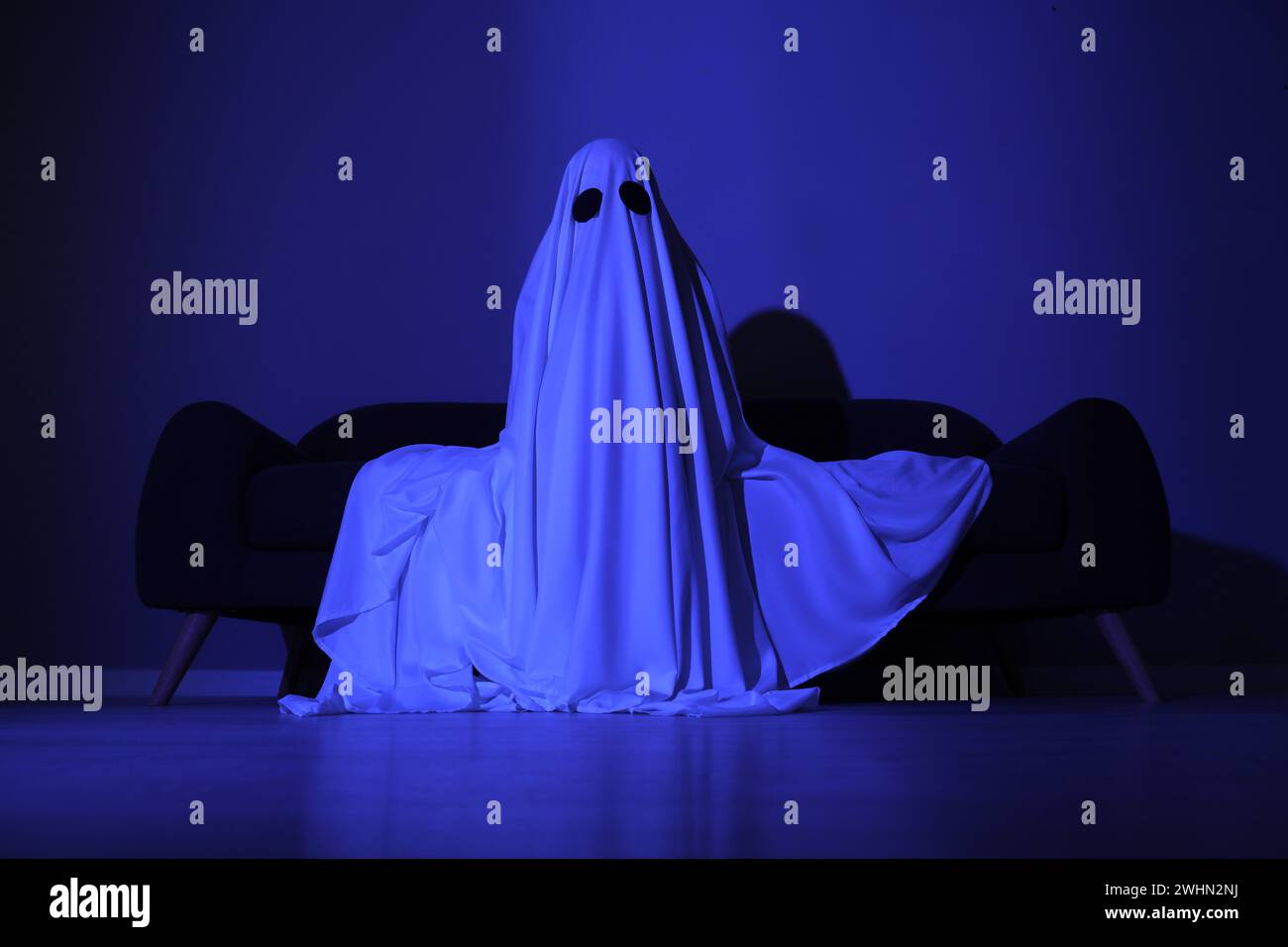 Creepy ghost. Woman covered with sheet sitting on sofa in blue light ...