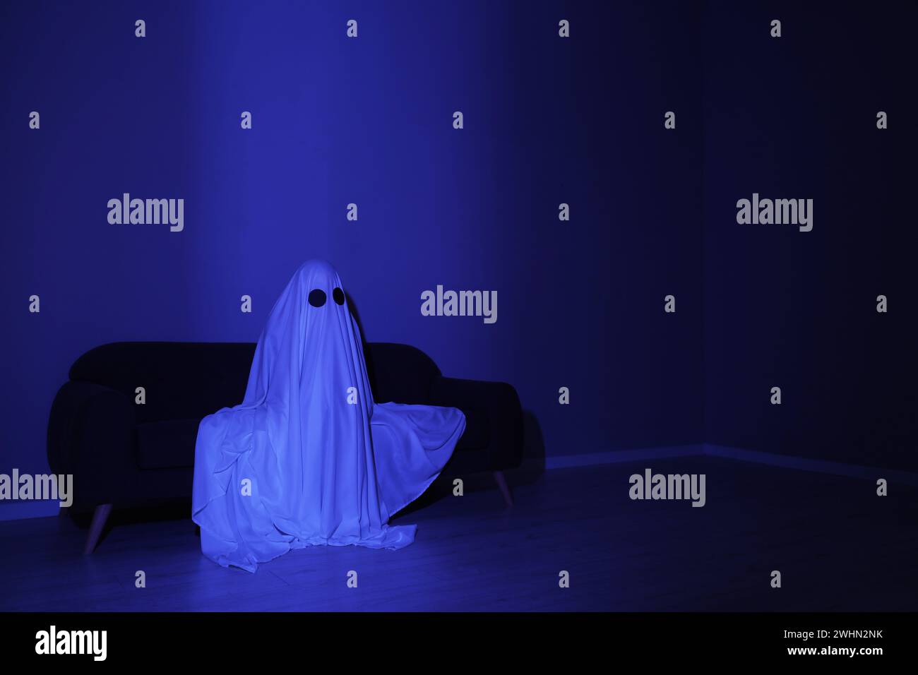 Creepy ghost. Woman covered with sheet sitting on sofa in blue light ...