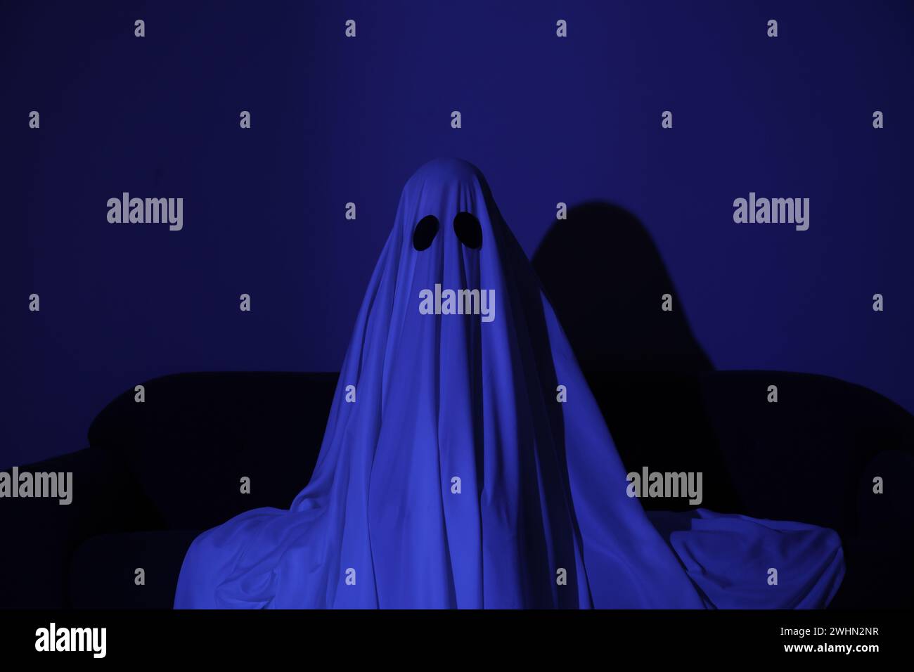 Creepy ghost. Woman covered with sheet sitting on sofa in blue light ...