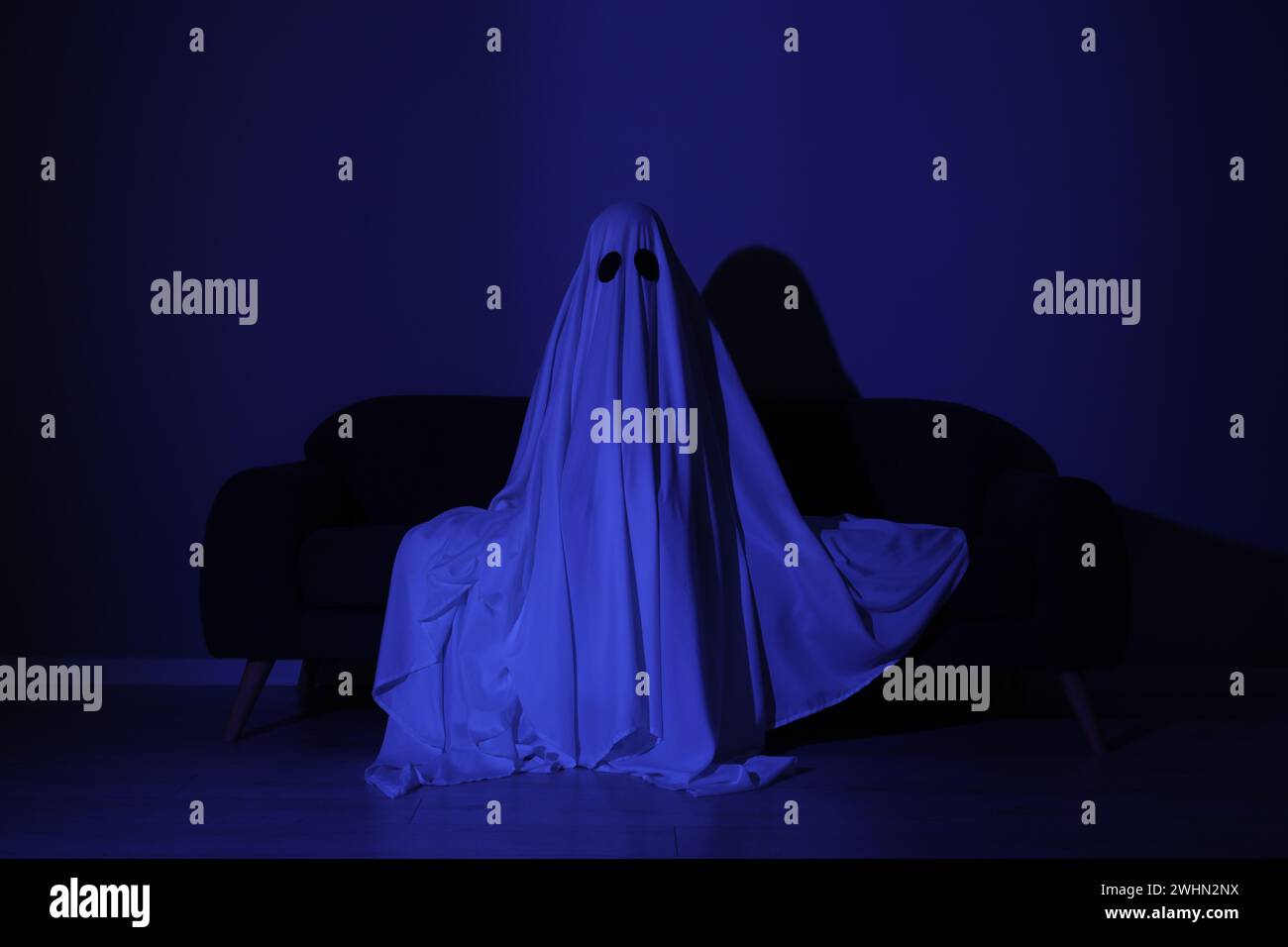 Creepy ghost. Woman covered with sheet sitting on sofa in blue light ...