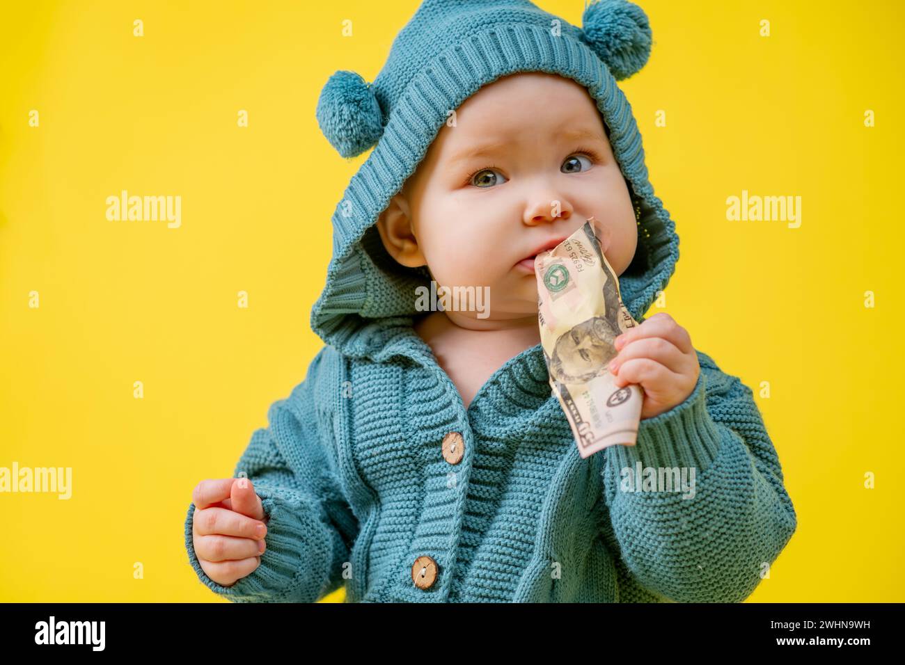 Cute kid chewing dollars on a yellow background. Baby in a blue knitted ...