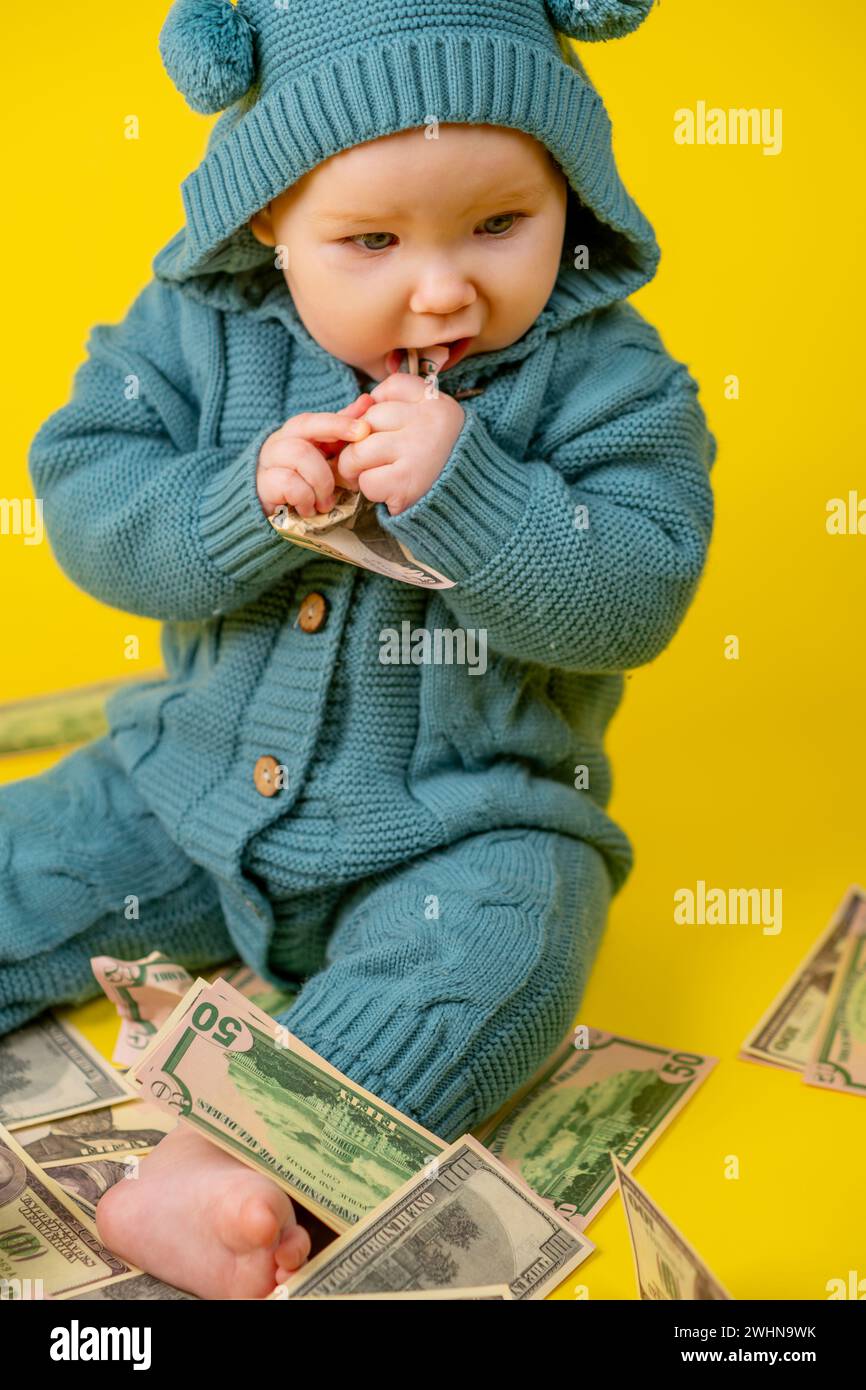 Cute kid chewing dollars on a yellow background. Baby in a blue knitted ...