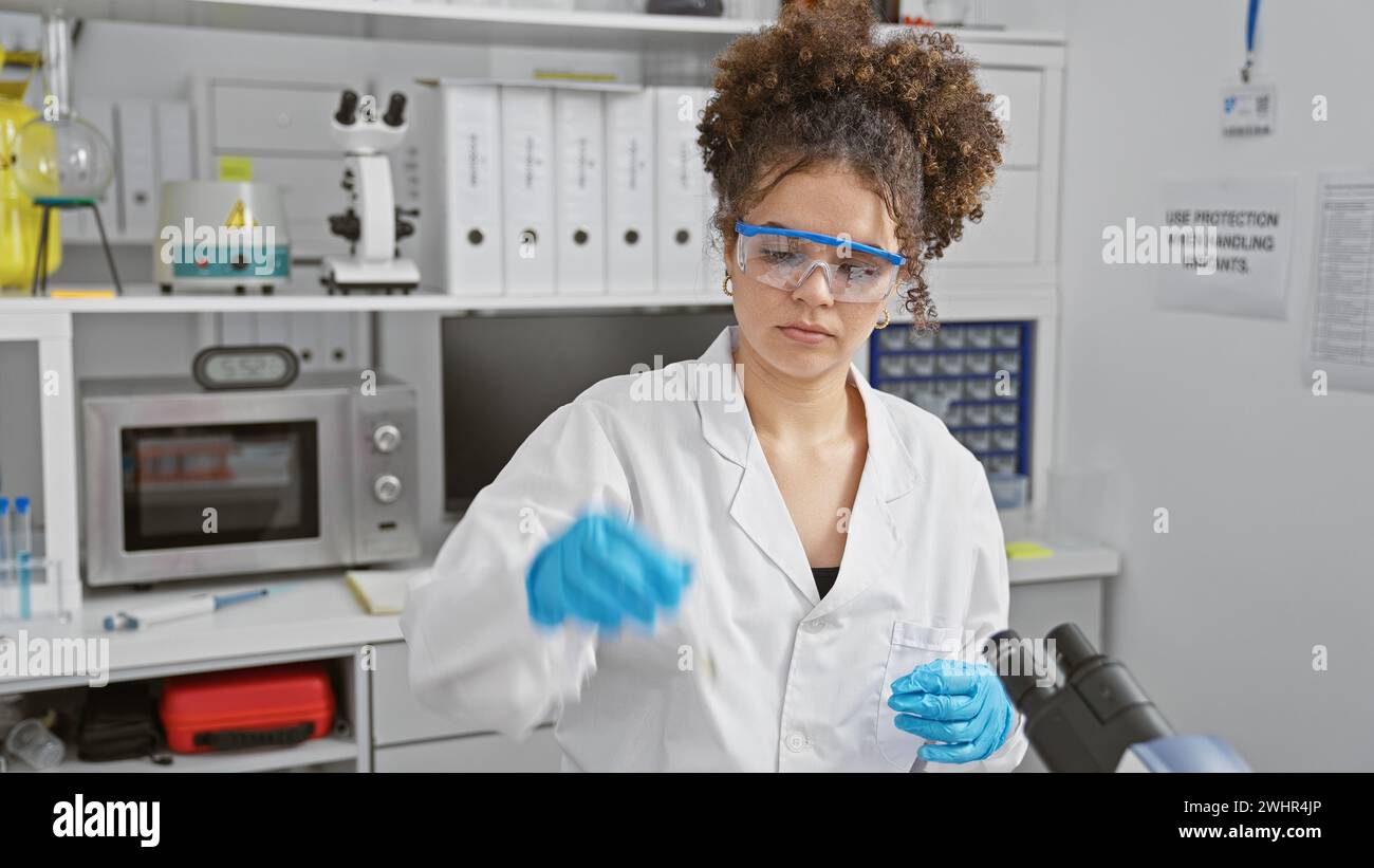 Hispanic woman with curly hair wearing safety goggles and lab coat in a ...