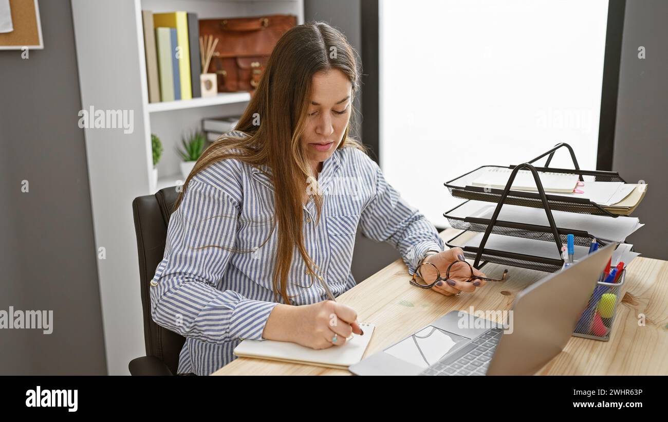 Focused woman writing notes in a modern office setting, with laptop and ...