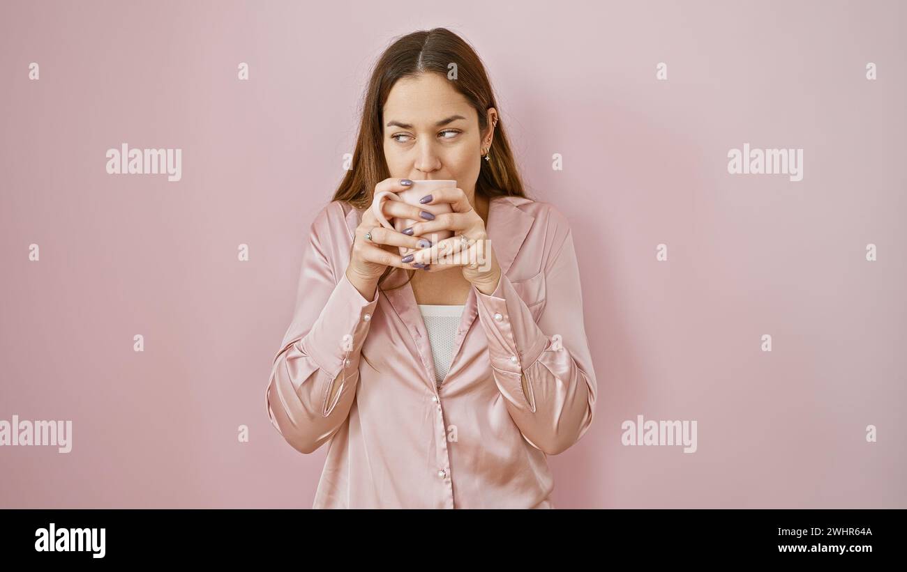 A contemplative young woman sips from a mug in a pink room, expressing ...