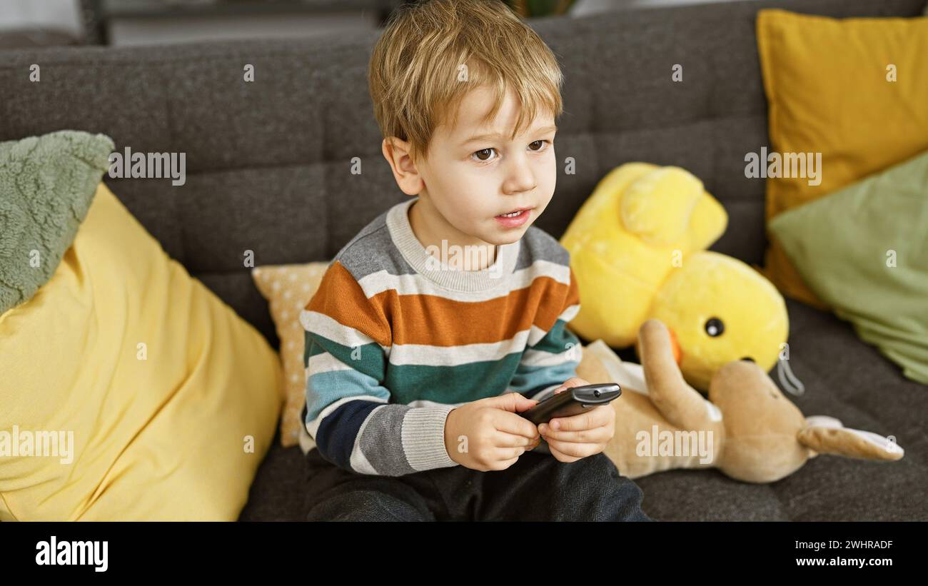 Blond toddler boy sitting indoors on a couch playing with a remote ...
