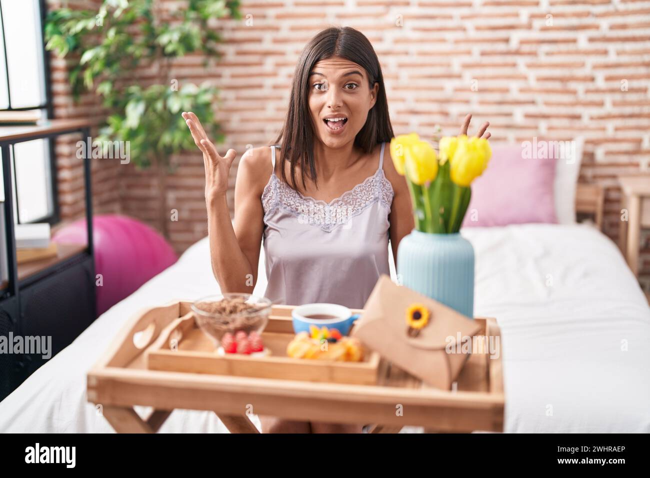 Brunette young woman eating breakfast sitting on the bed celebrating victory with happy smile ...