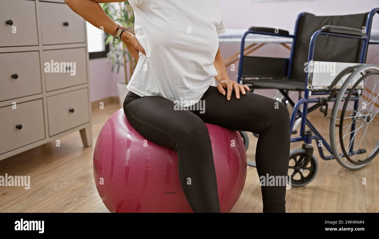 A middle-aged woman with back pain sits on a therapy ball in a ...