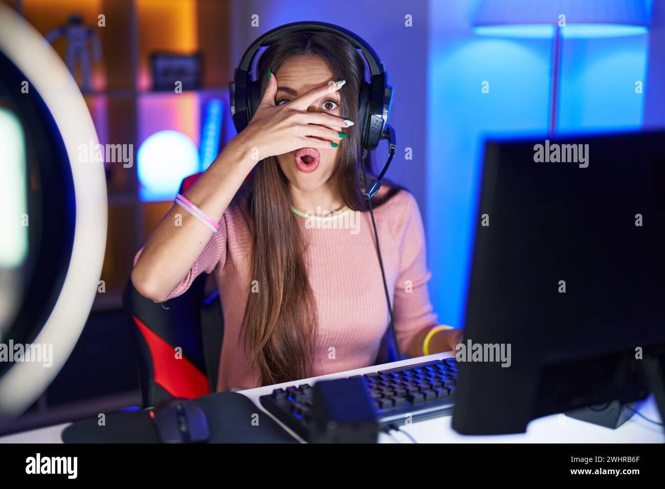 Young hispanic woman playing video games peeking in shock covering face ...