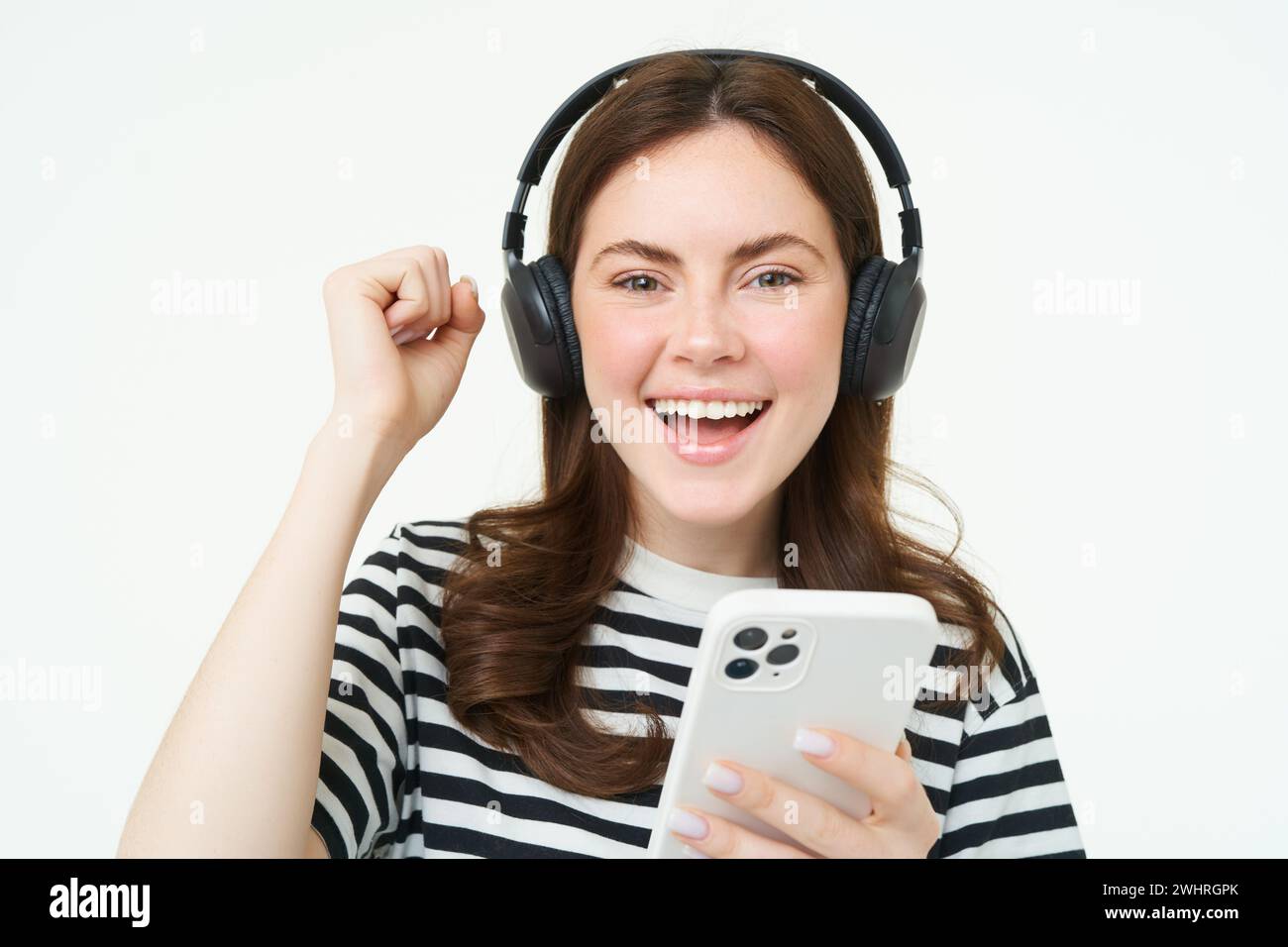 Enthusiastic girl in headphones, holding smartphone, smiling and ...