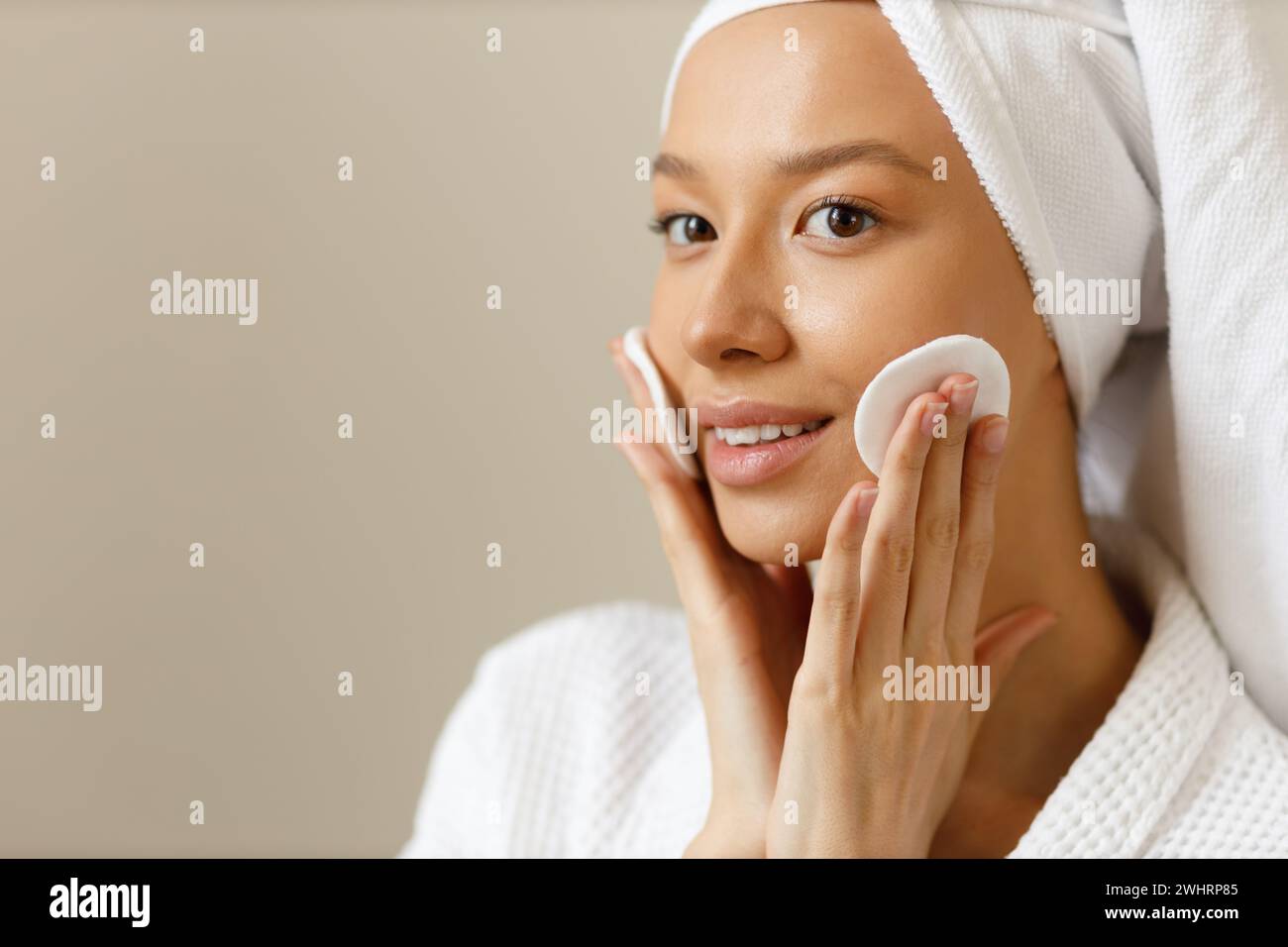 Pretty woman in robe and towel on her head wipes the skin of her face with cotton pads ...