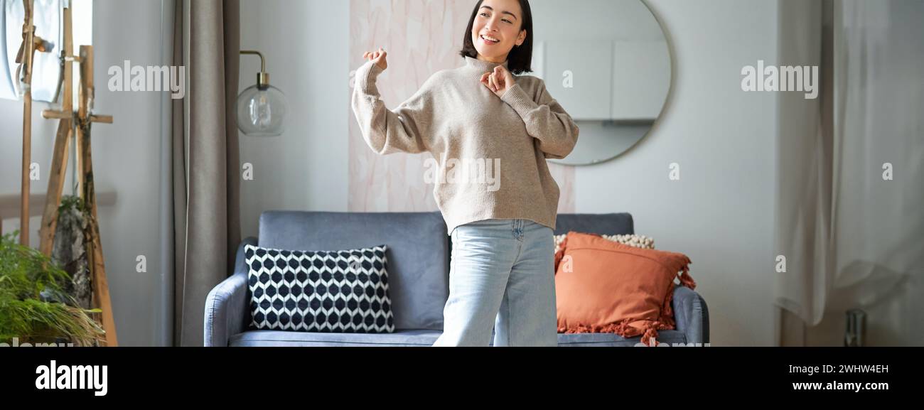 Enthusiastic asian woman dancing, feeling free and upbeat at her home ...