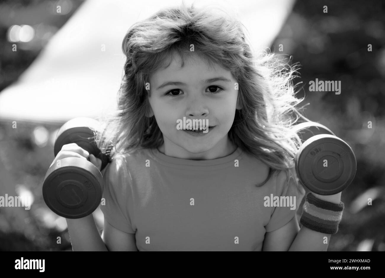 Cute child boy pumping up arm muscles with dumbbell outdoor in summer ...