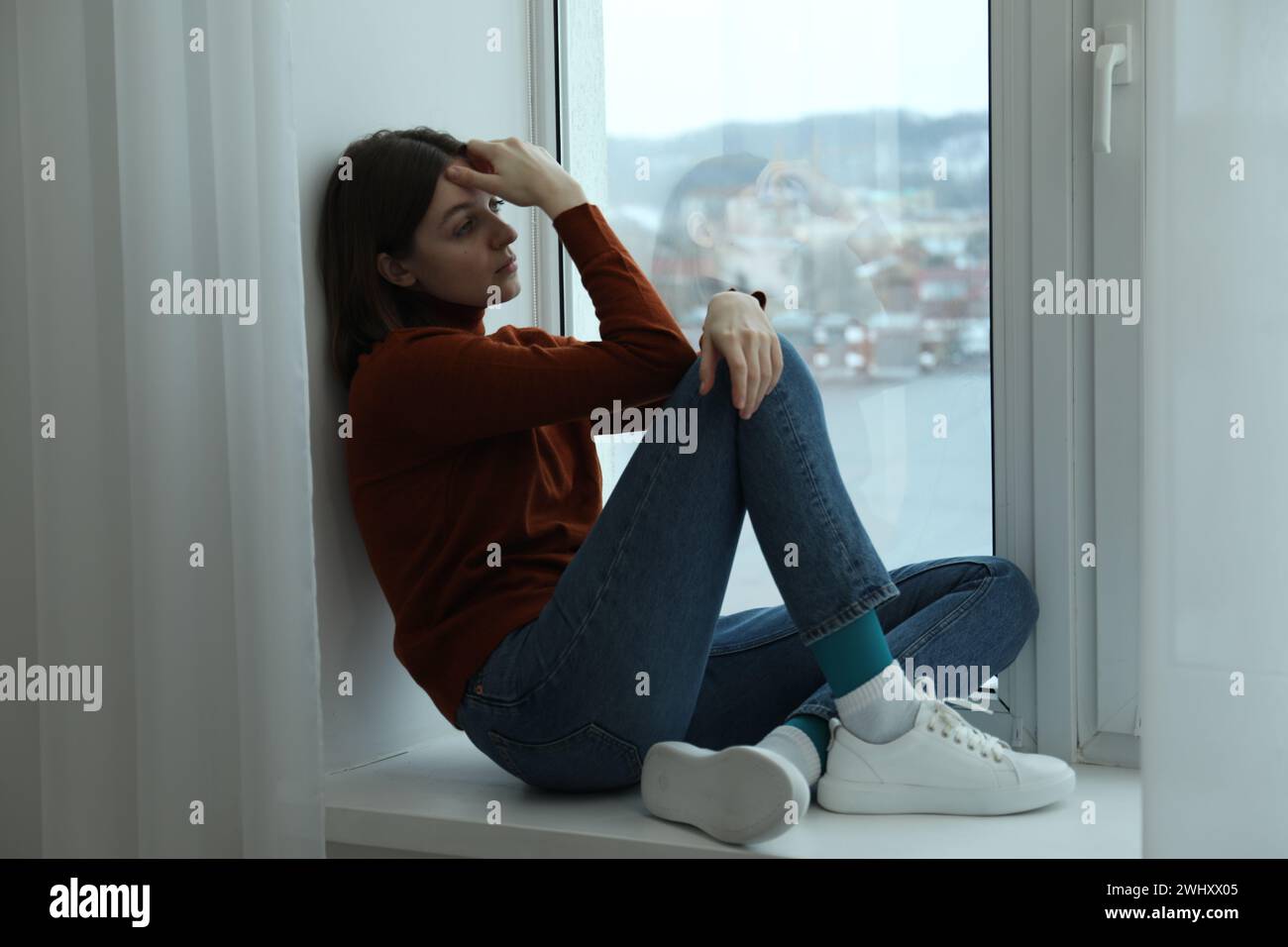 Sad young woman sitting on windowsill near window at home Stock Photo ...