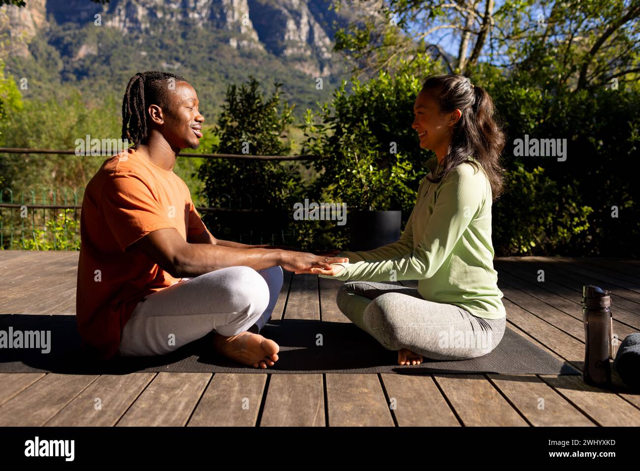 Happy diverse couple practicing yoga meditation sitting holding hands on deck in sunny garden ...