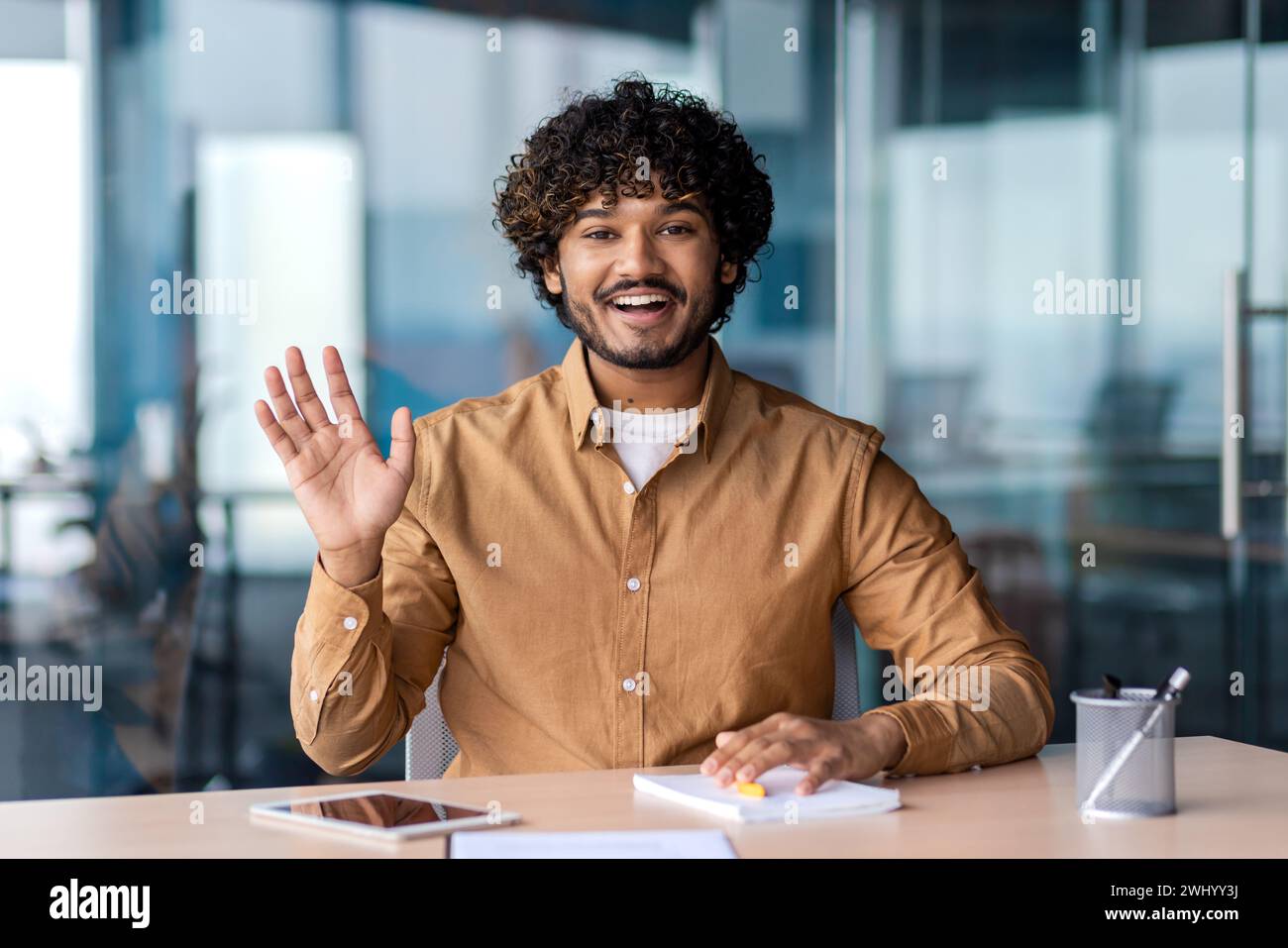 Smiling hispanic employee making hello gesture while keeping hand on ...