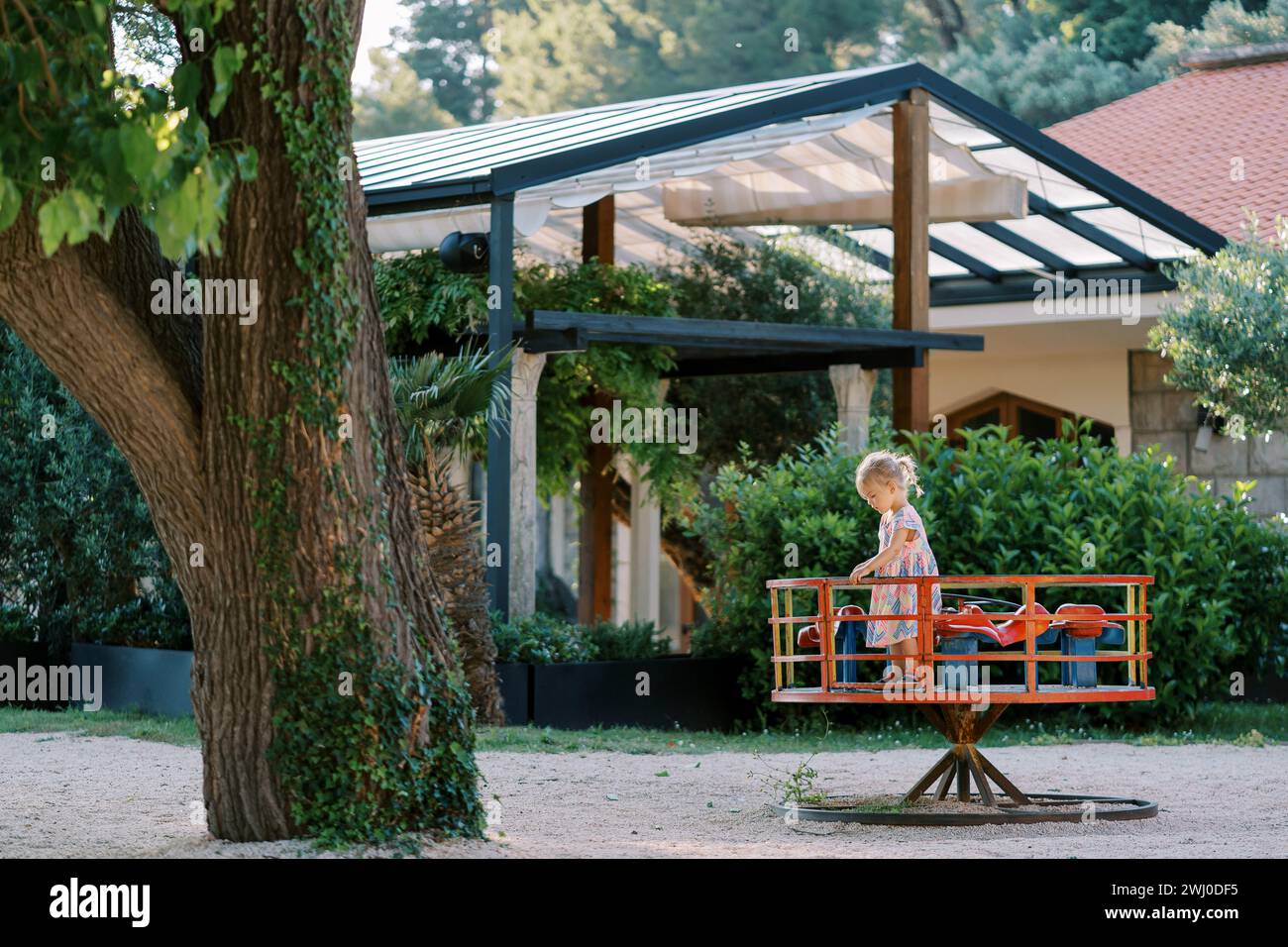 Little girl stands on a carousel in a green garden. Side view Stock ...