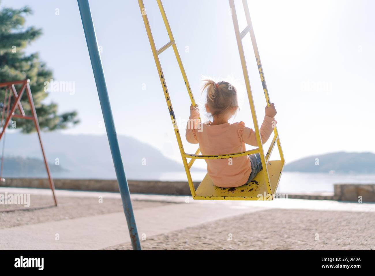 Little girl swings on a swing by the sea in bright sunlight. Back view Stock Photo - Alamy