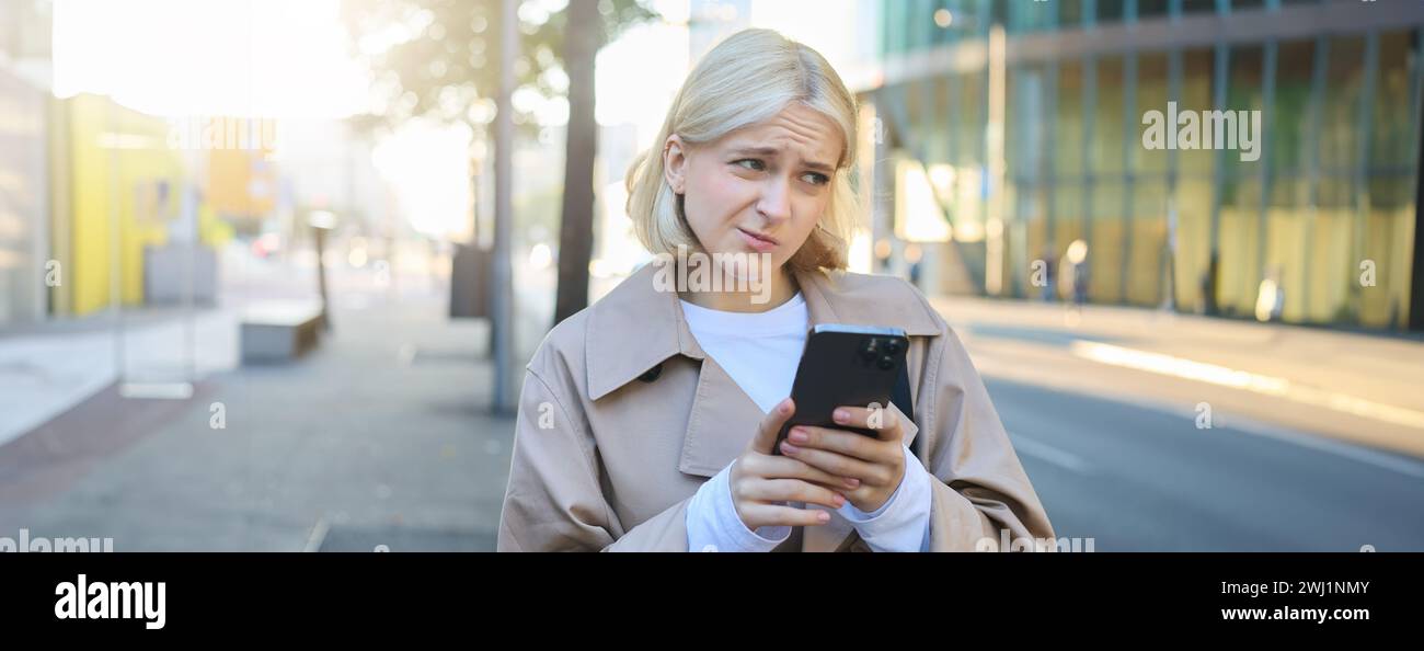Portrait of young blond woman standing on street, has unsure, doubtful ...