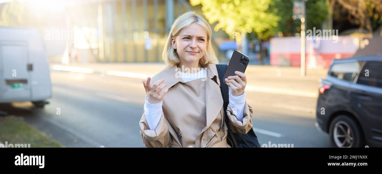 Portrait of confused blonde woman on street, carries backpack, holding ...