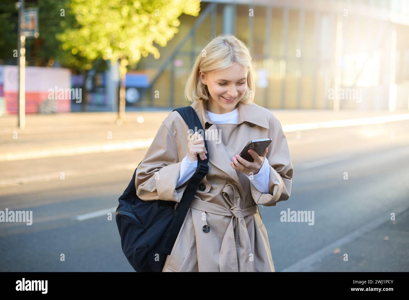Smiling blonde woman in trench coat, standing on street, using mobile ...