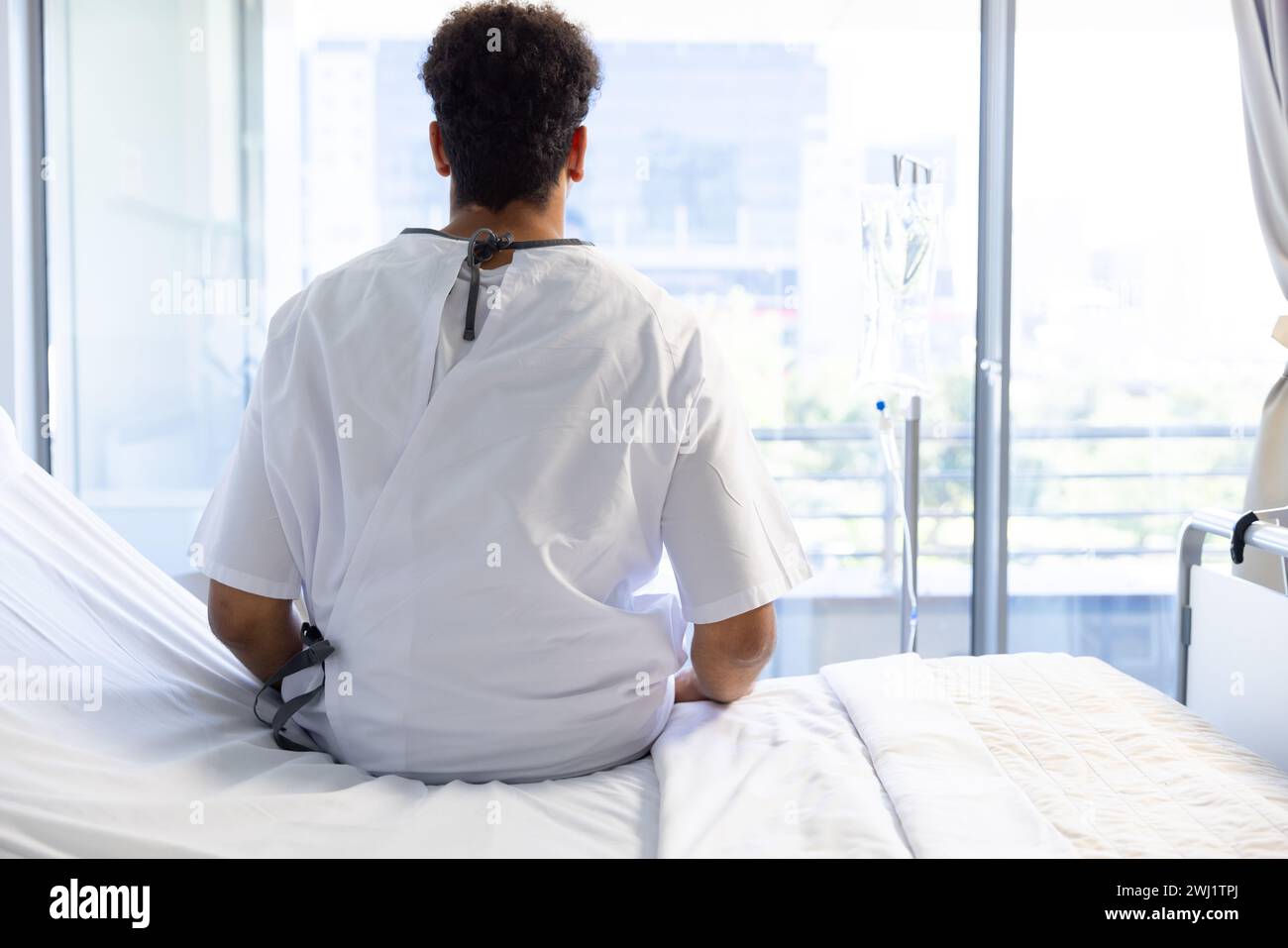 Back of biracial male patient sitting on bed in sunny hospital room ...