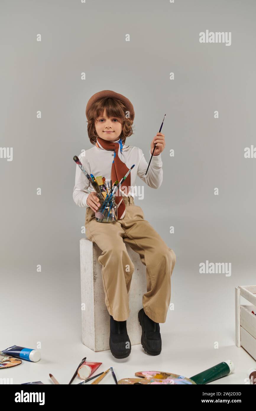 stylish artist sitting on white block with paint brushes, boy in beret ...