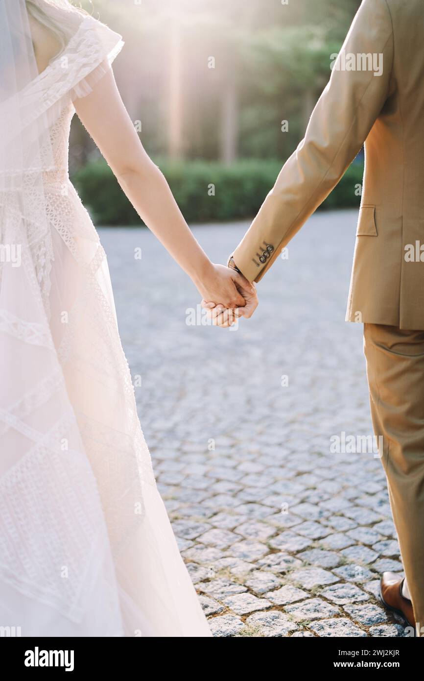 Bride and groom walk holding hands along the paving stones in the park ...