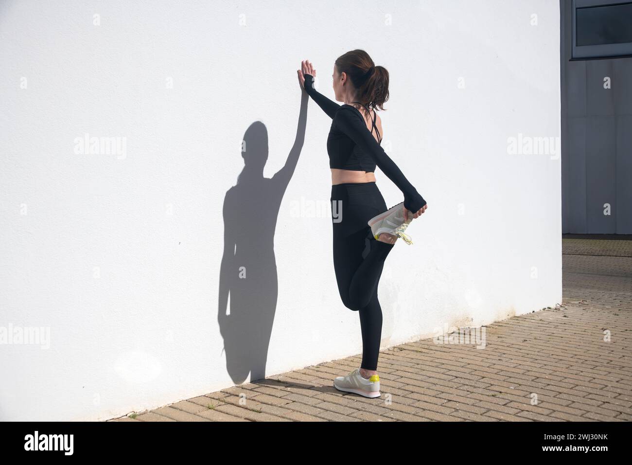 Sporty woman doing warm up exercises, leg stretch against a white wall ...