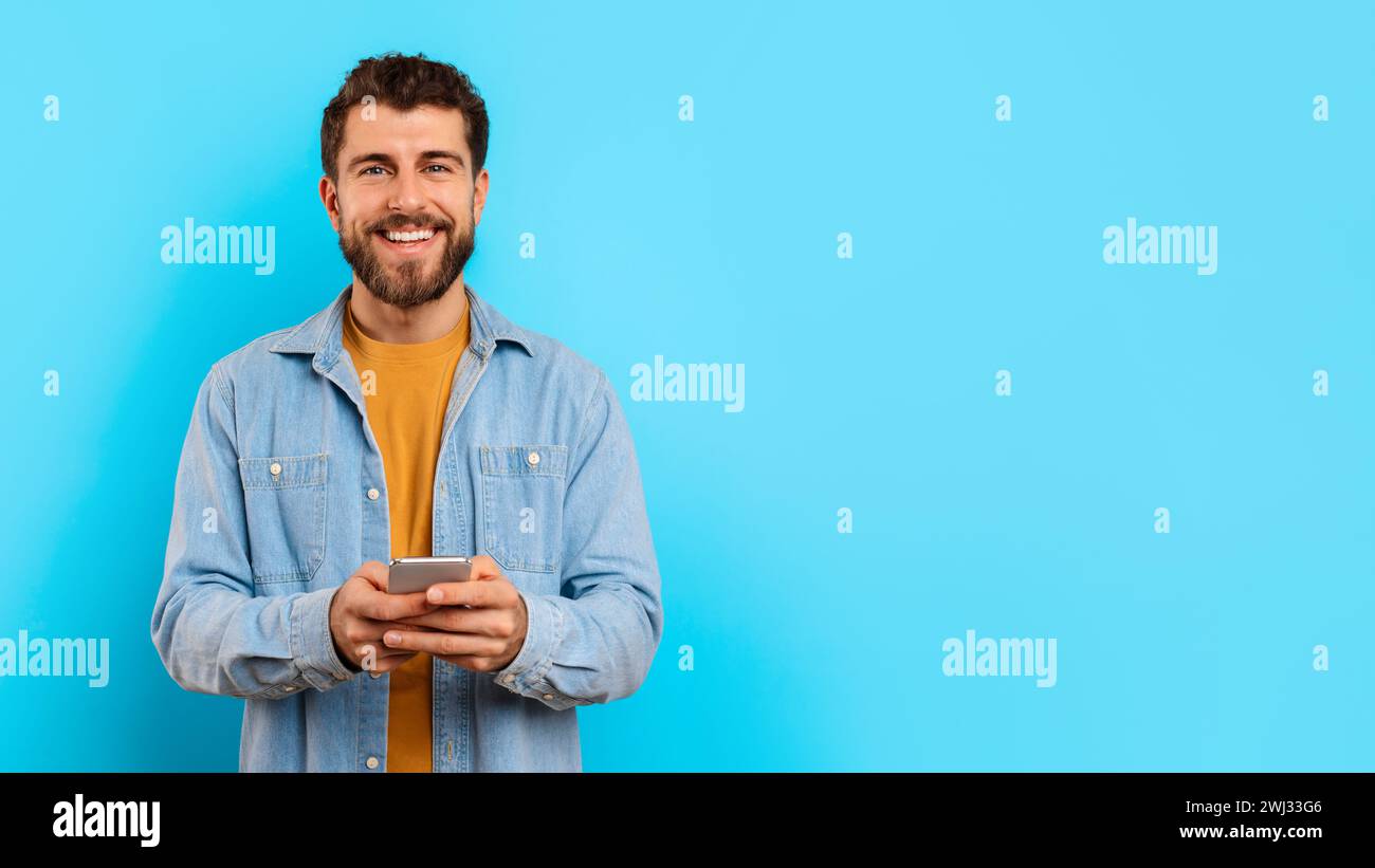 Guy holding smartphone scrolling and texting against blue studio ...
