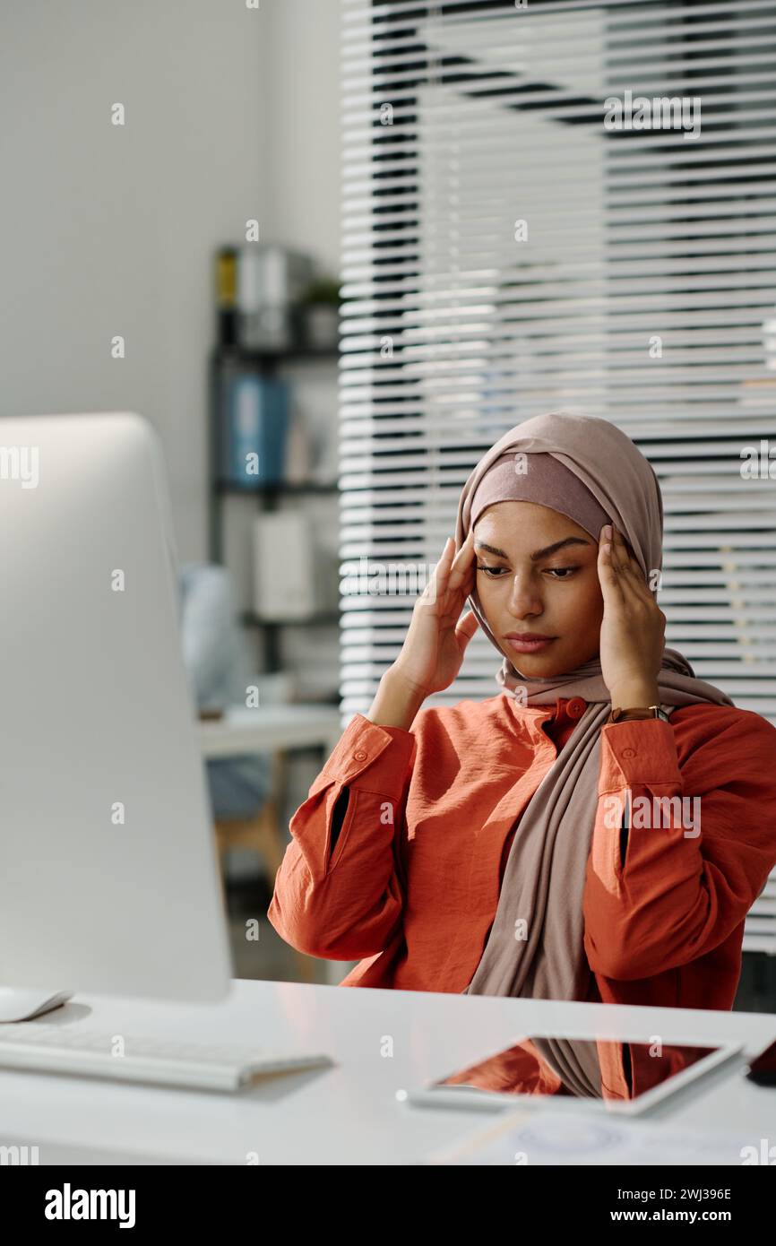 Young tired female manager touching her head while sitting by workplace ...