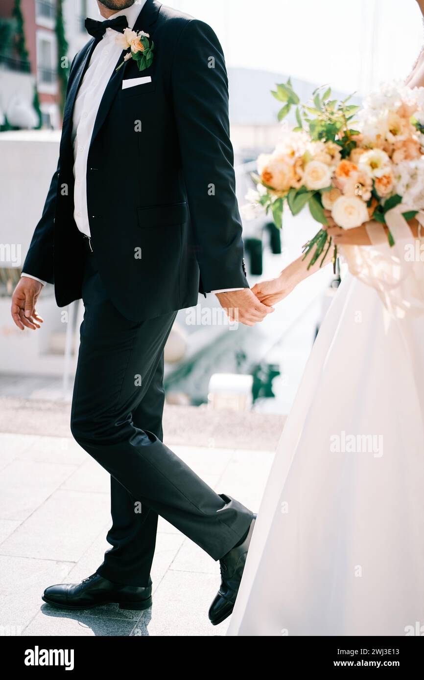 Bride and groom with a bouquet walk holding hands along the sea ...