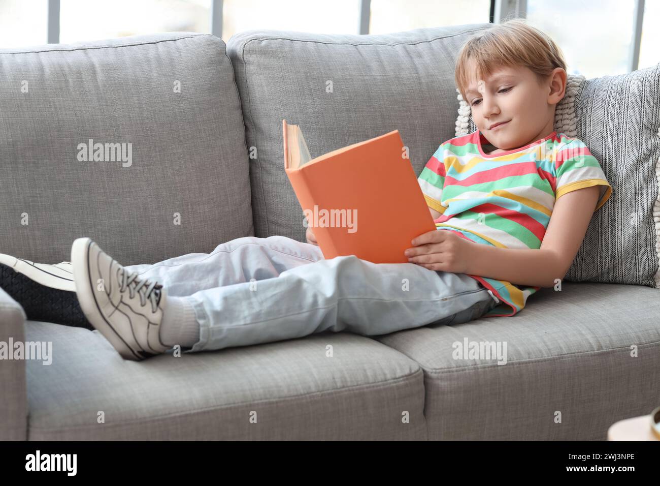 Cute little boy reading book while lying on sofa Stock Photo - Alamy
