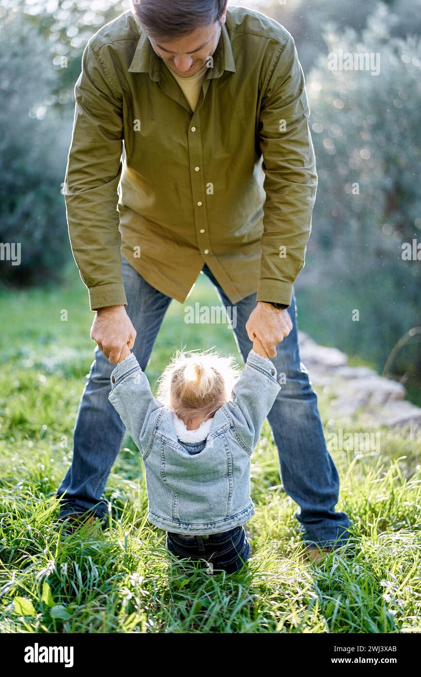 Dad stands over a little girl sitting on the lawn and holds her hands ...