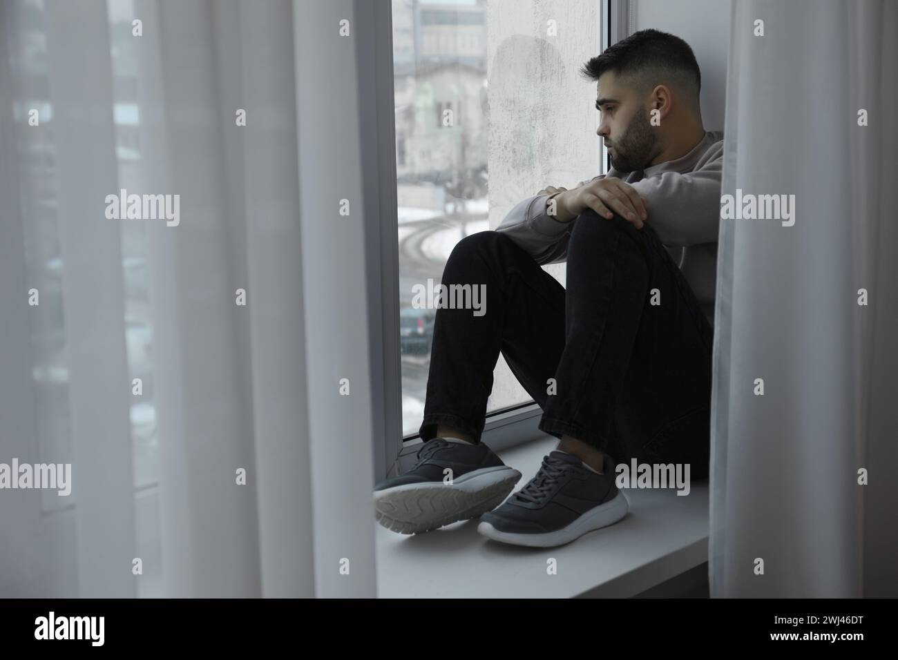 Sad man sitting on sill and looking at window indoors Stock Photo - Alamy