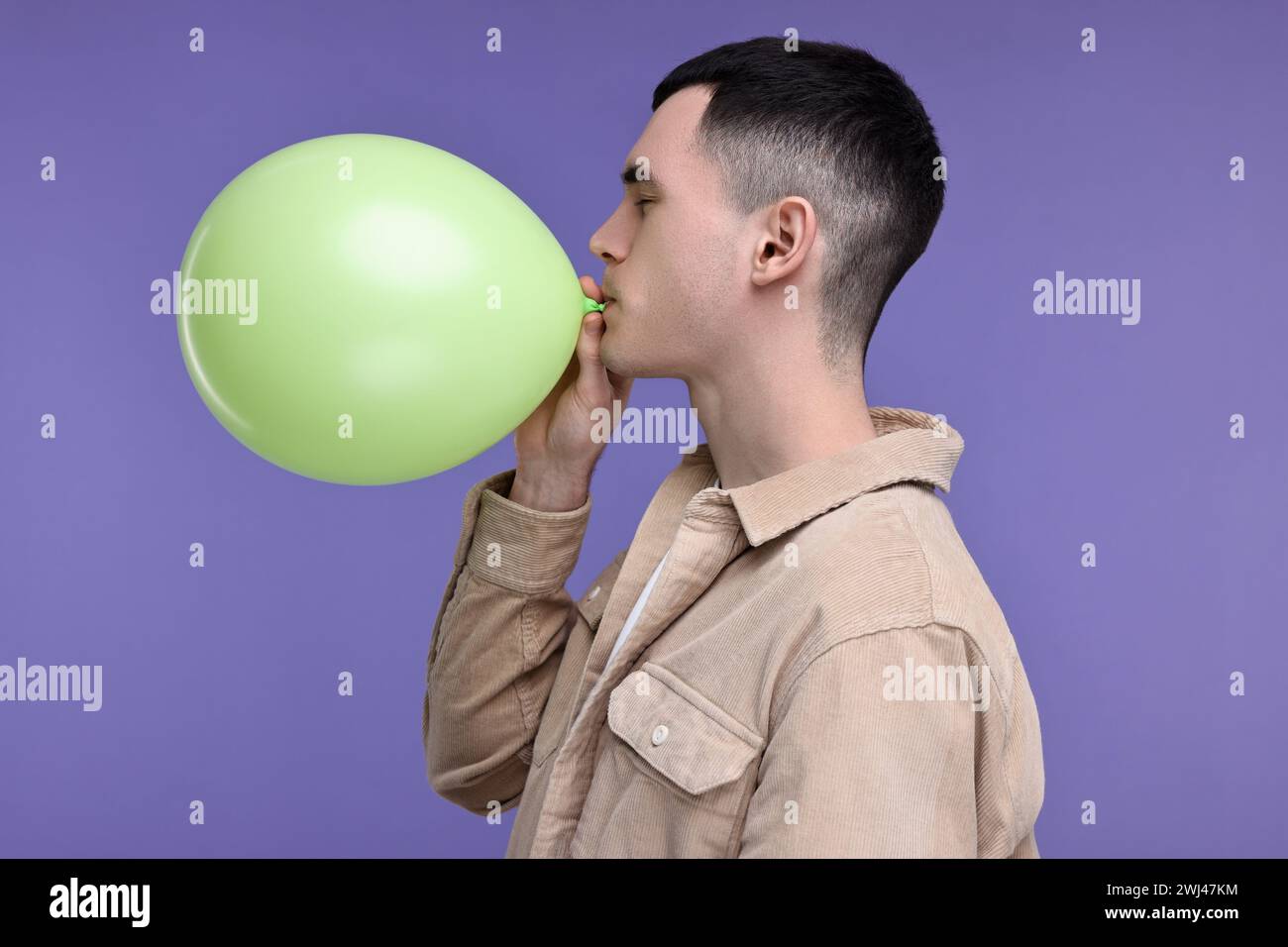 Young man inflating light green balloon on purple background Stock ...