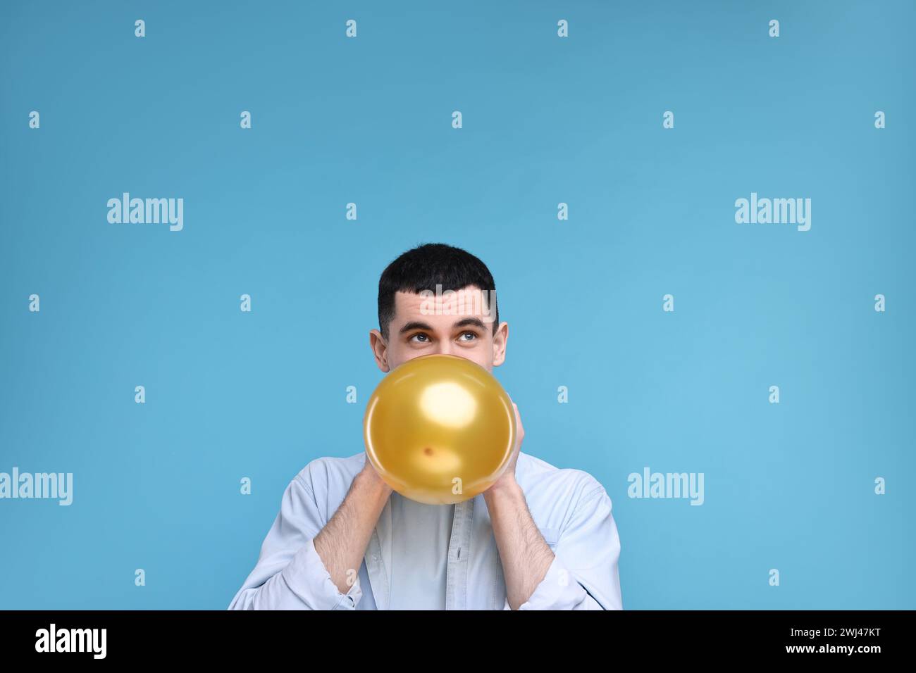Young man inflating golden balloon on light blue background. Space for ...