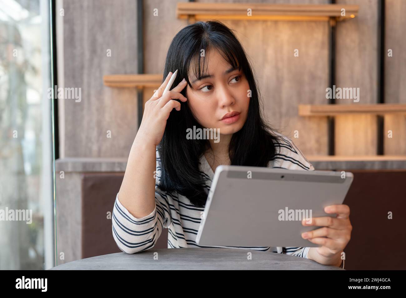 A confused, serious Asian woman, working remotely at a cafe, is using ...