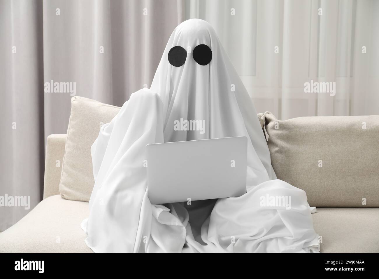 Creepy ghost. Person covered with white sheet using laptop on sofa at ...