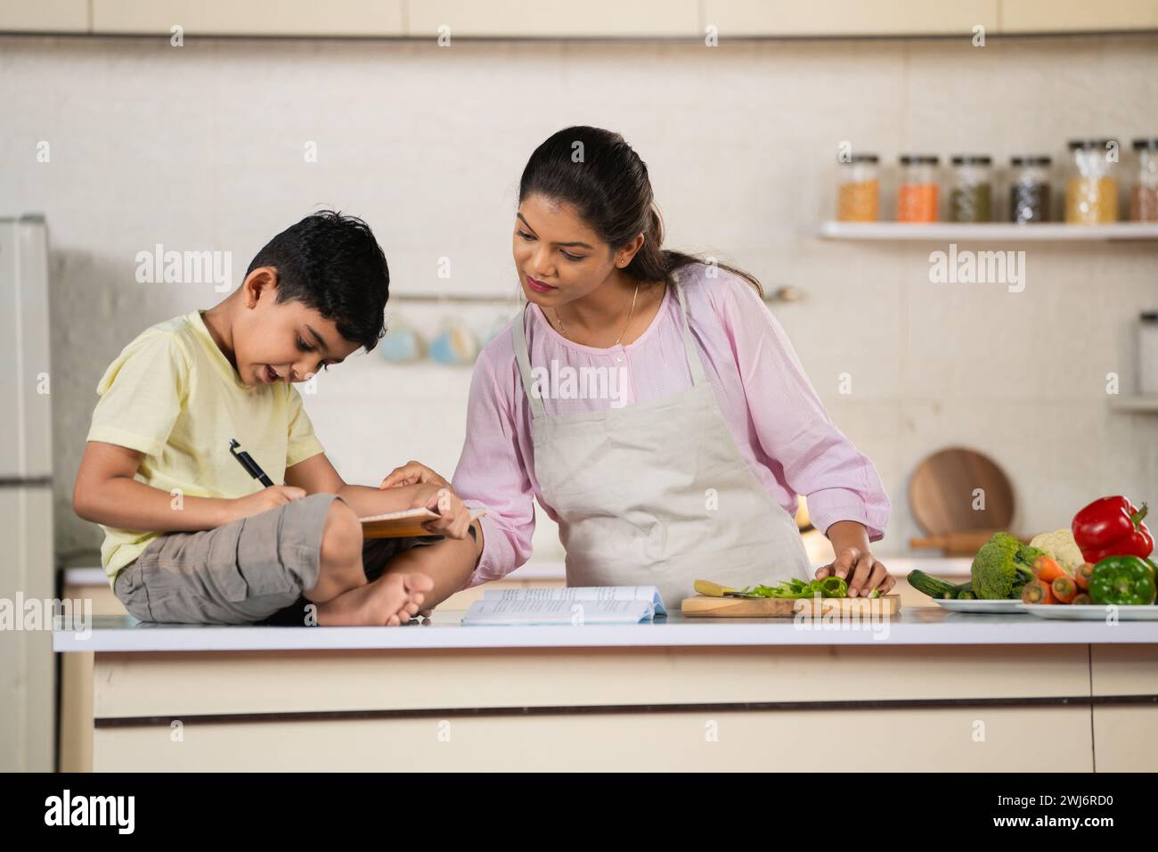 Indian working mother at kitchen helping her kid or child to do ...