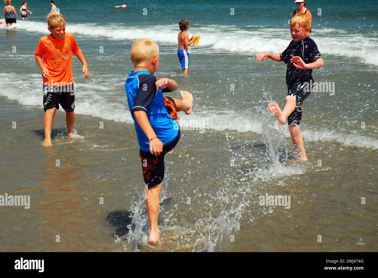 Young friends play in the ocean waves at the beach, kicking water at ...