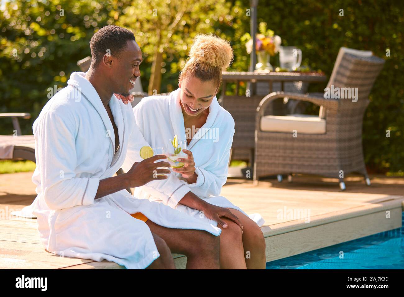 Couple Wearing Robes Outdoors Sitting With Drinks By Swimming Pool On ...