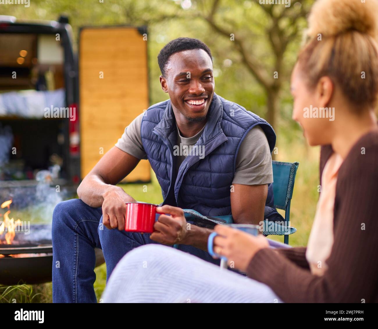 Couple Camping In Countryside With RV Sitting In Chairs Drinking Coffee ...