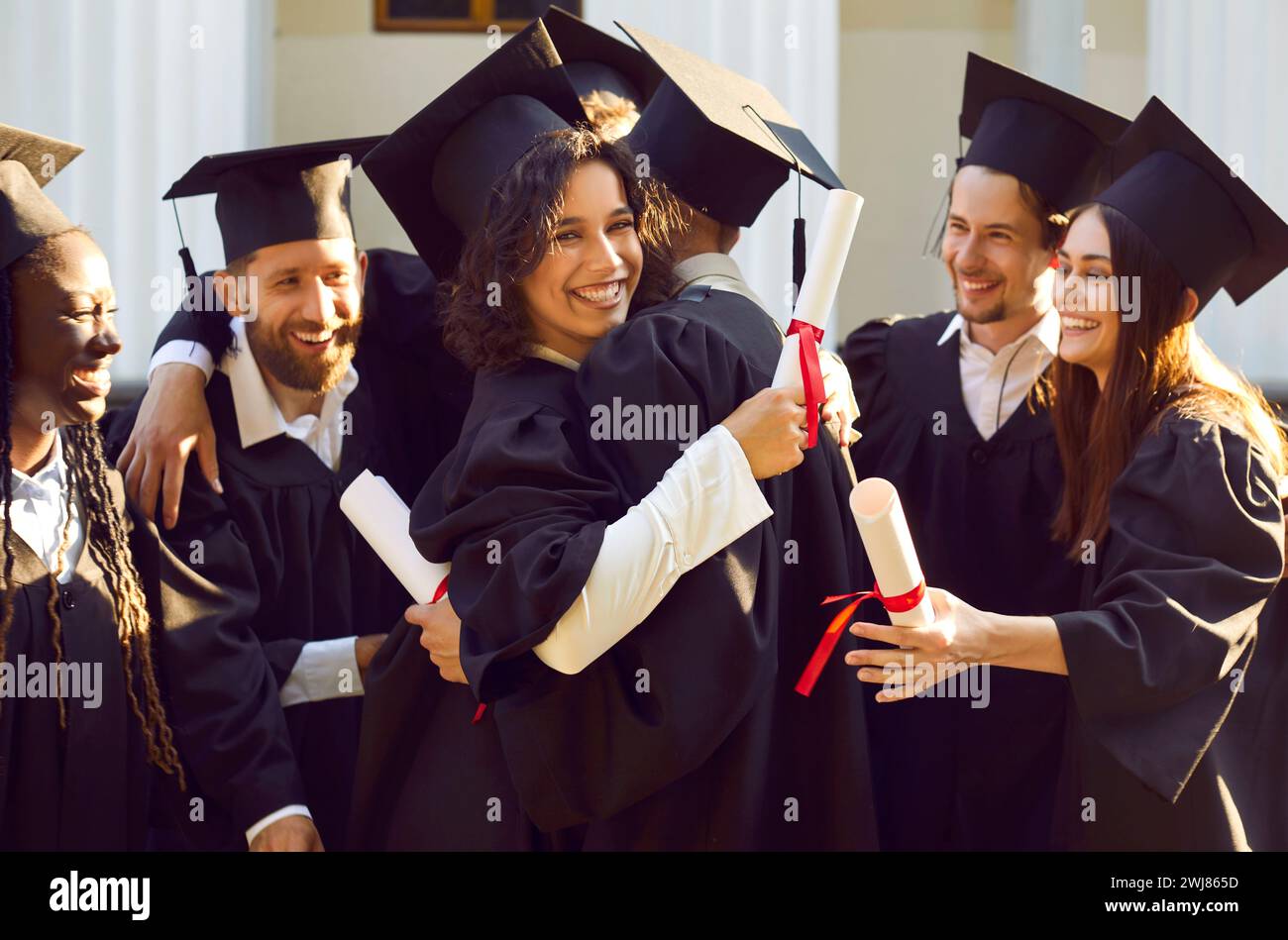 Happy diverse graduates hugging outdoors holding diplomas and ...