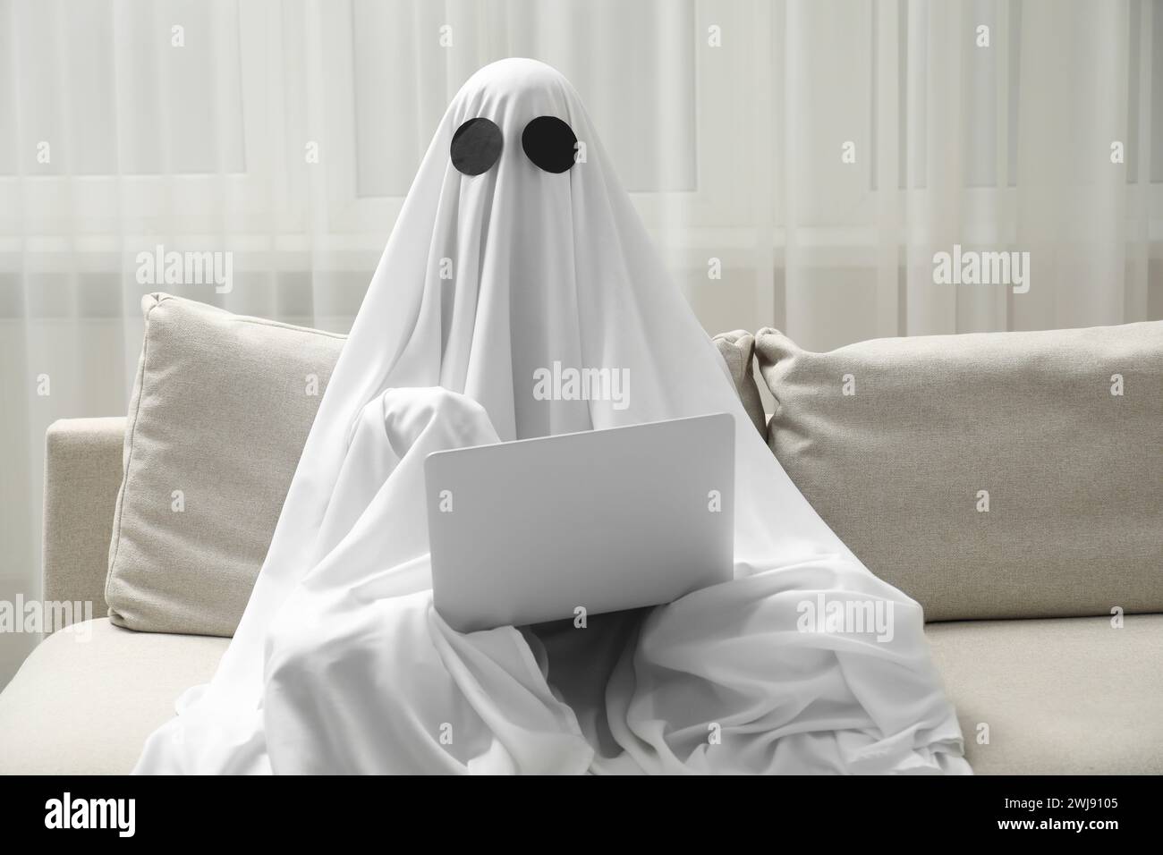 Creepy ghost. Person covered with white sheet using laptop on sofa at ...