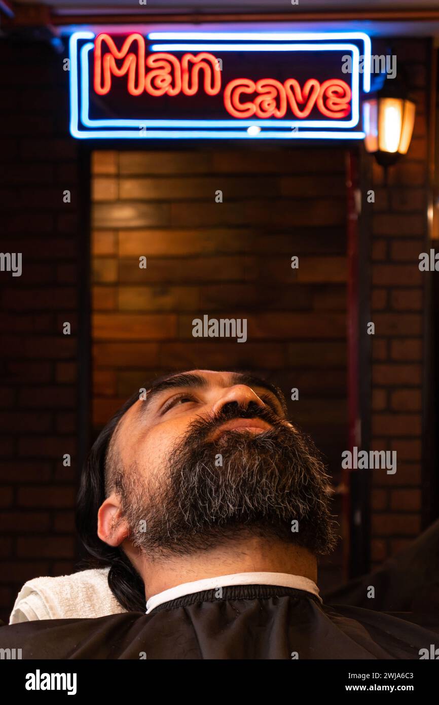 Bearded client in barber cape sitting on chair and looking up during ...