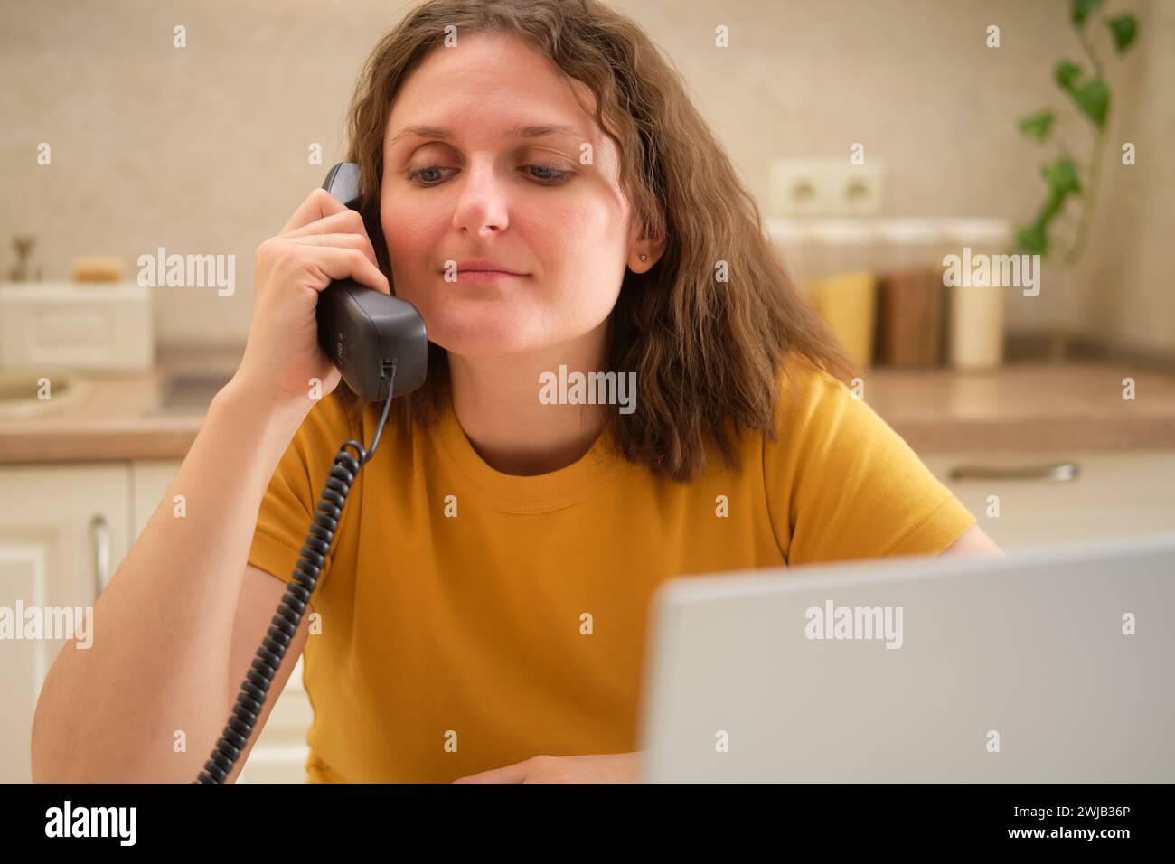 A woman with a laptop is talking on a landline phone at a table in a ...