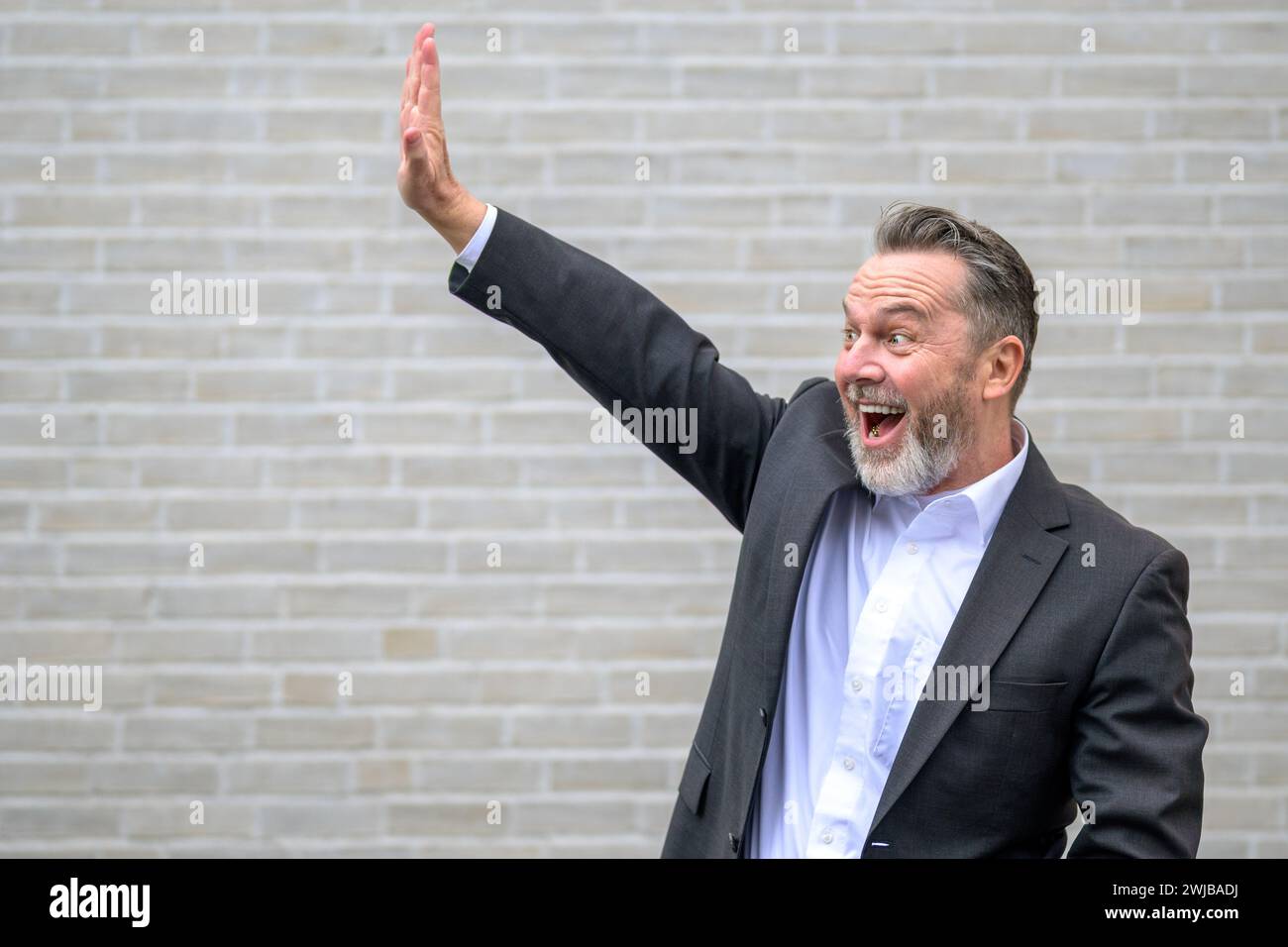 Happy sixty year old gray man in front of a white brick wall waving to ...