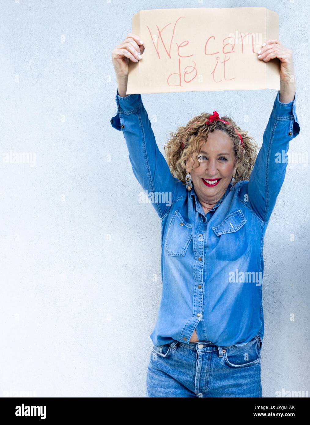 A smiling woman in blue denim holds up a 'We can do it' sign, embodying ...